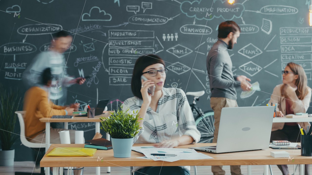 Zoom-in time lapse of young woman working in open space office with people moving around